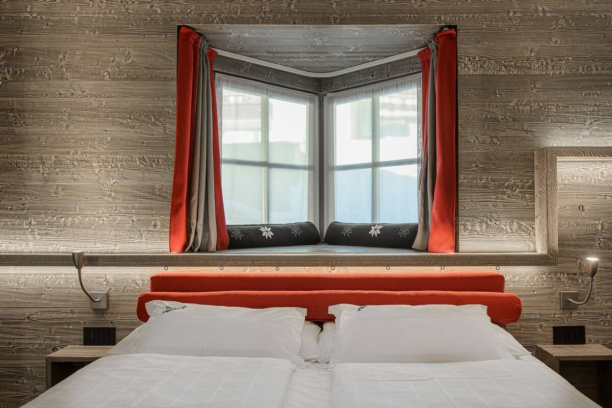 Quarto de canto com parede de madeira, janela com cortinas vermelhas, cabeceira vermelha e mesa de cabeceira com abajur.