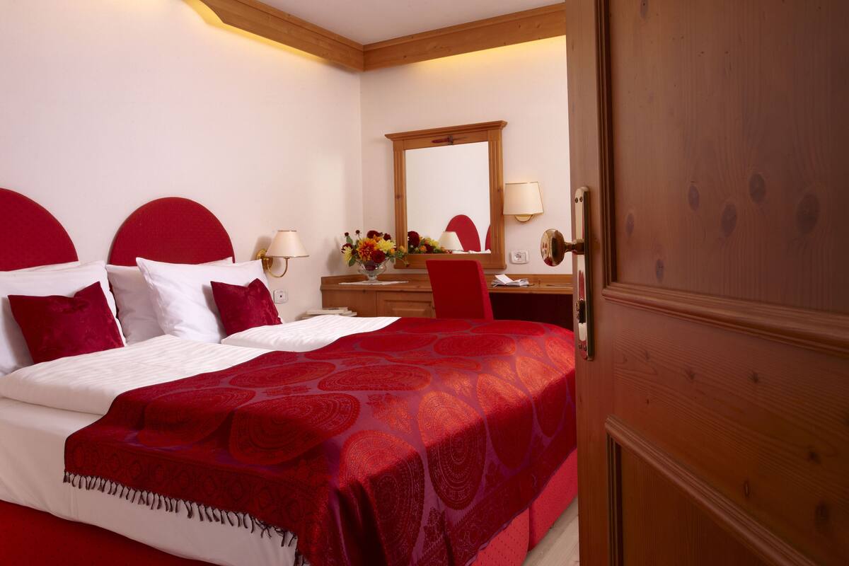 Quarto de hotel com uma cama grande, cabeceira vermelha, roupa de cama branca, uma escrivaninha de madeira com espelho e um cobertor estampado em vermelho.