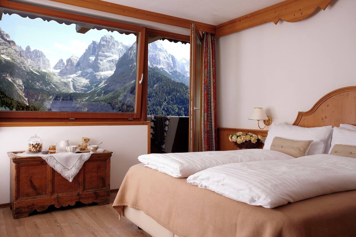 Quarto com cama, penteadeira de madeira, janela com vista para as montanhas e abajur na mesa de cabeceira.