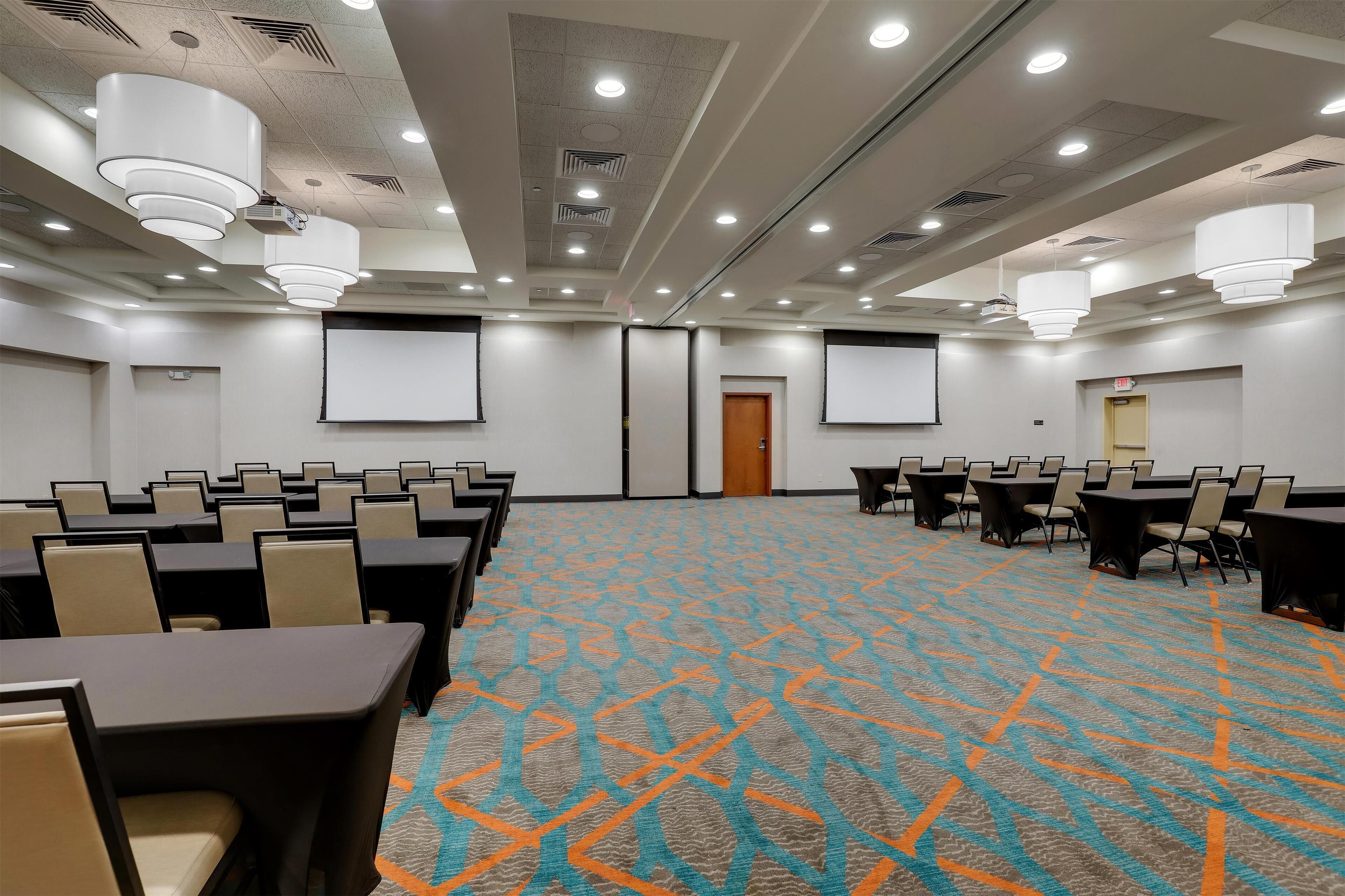 Sala de conferências com carpete estampado, várias mesas, cadeiras e um projetor.