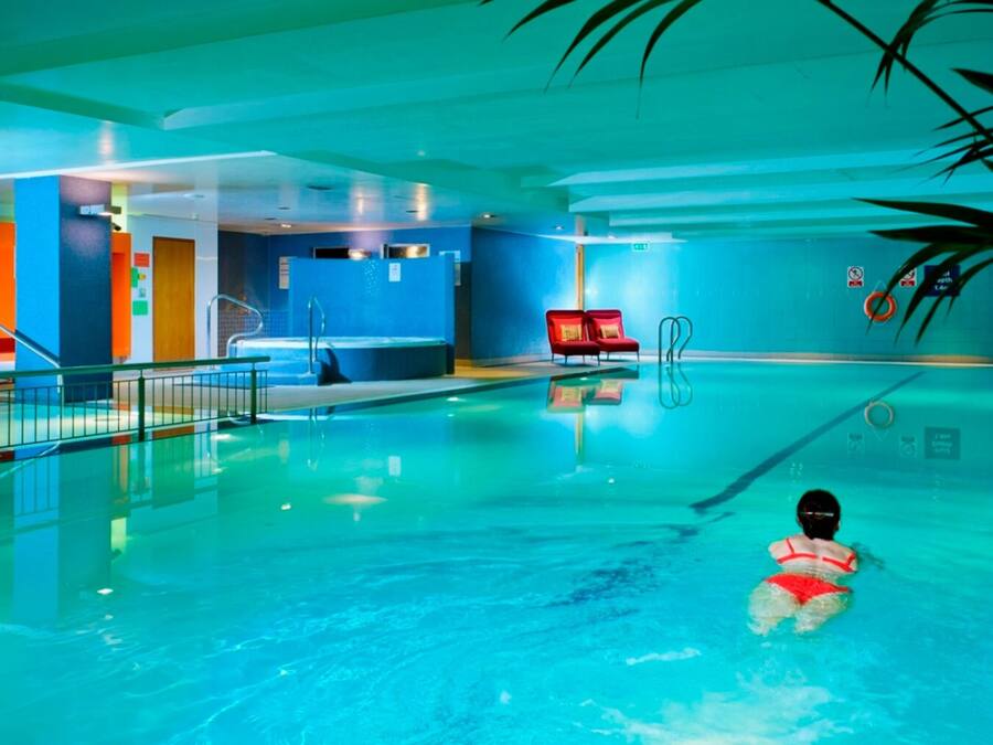 Piscina interior com uma criança nadando, cercada por paredes azuis e uma cadeira vermelha.
