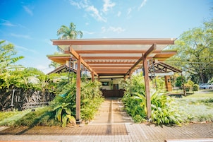 Uma galeria coberta leva à entrada de um hotel, com vegetação exuberante e céu claro.