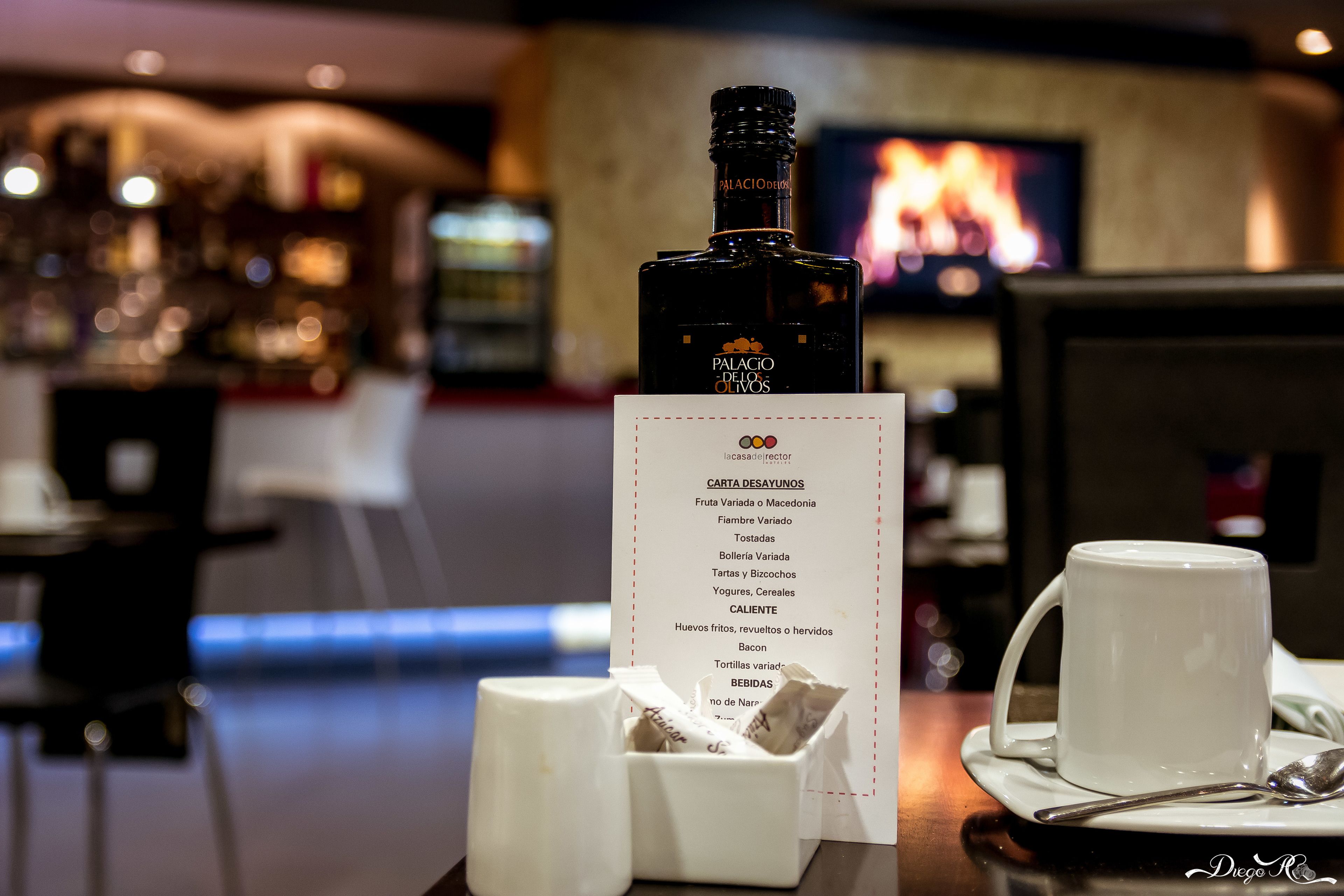 Uma área de bar com uma garrafa de Palacio del Olivo, um menu listando itens de café da manhã e uma xícara branca em um pires.