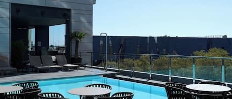 Uma área de piscina no terraço com espreguiçadeiras e mesas, vista desobstruída do horizonte urbano e fachada de edifício moderna.