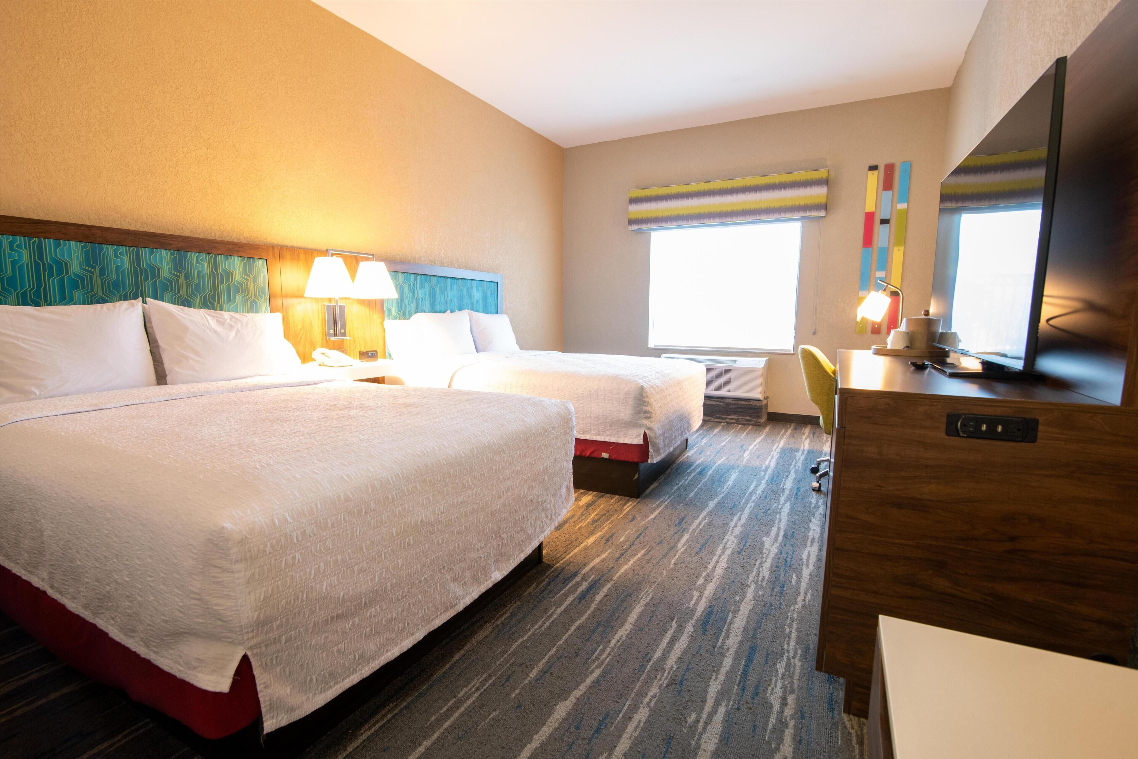 Quarto de hotel com duas camas, uma escrivaninha, uma cadeira, uma televisão e uma janela com persianas.