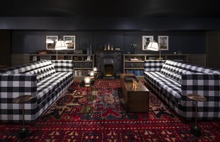 Uma sala de estar com decoração escura, sofás estampados em xadrez, lareira, estantes de livros e tapete com padrão.