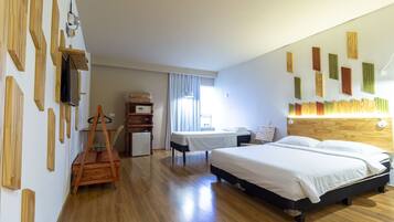 Quarto de hotel moderno com piso de madeira, cama com roupa de cama branca, escrivaninha com cadeira e uma pequena mesa de trabalho com luminária.