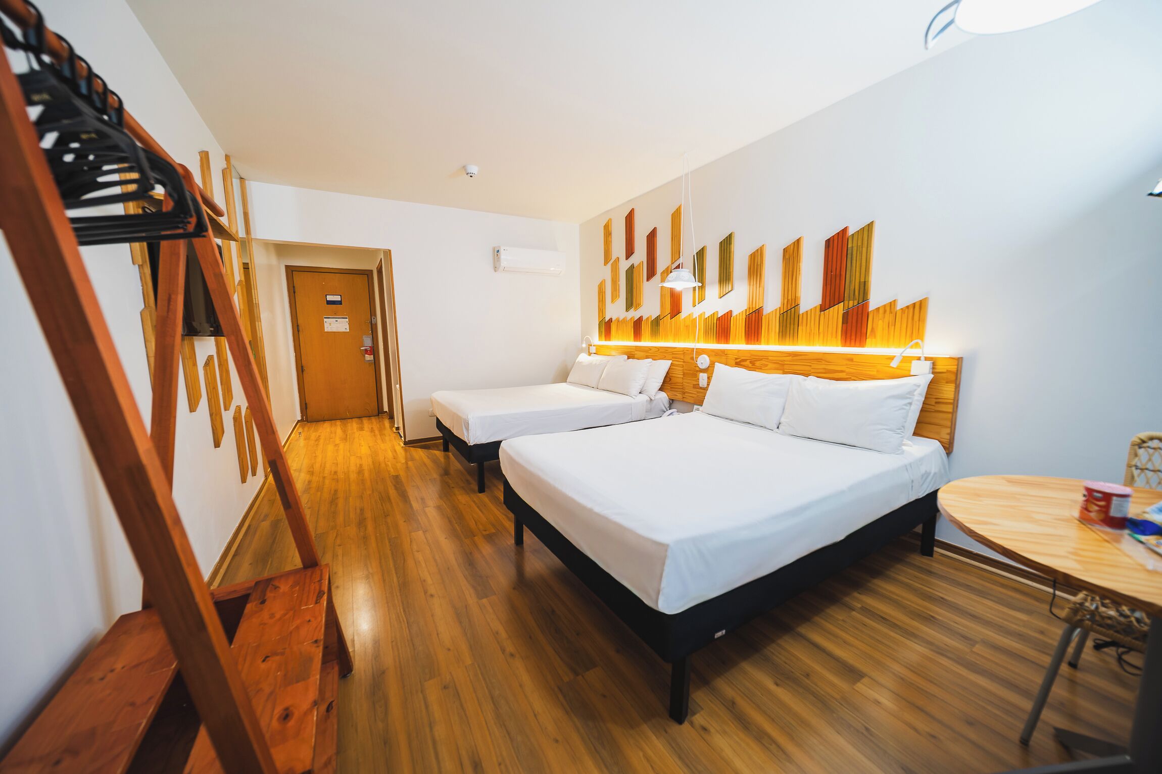 Quarto de hotel com duas camas de solteiro, piso de madeira, uma mesa redonda pequena e luminária fixada na parede.