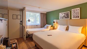 Quarto de hotel com duas camas, uma mesa de cabeceira com luminária, uma janela com cortinas e um quadro pendurado na parede.