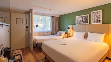 Quarto de hotel com duas camas, uma mesa de cabeceira com luminária, uma janela com cortinas e um quadro pendurado na parede.