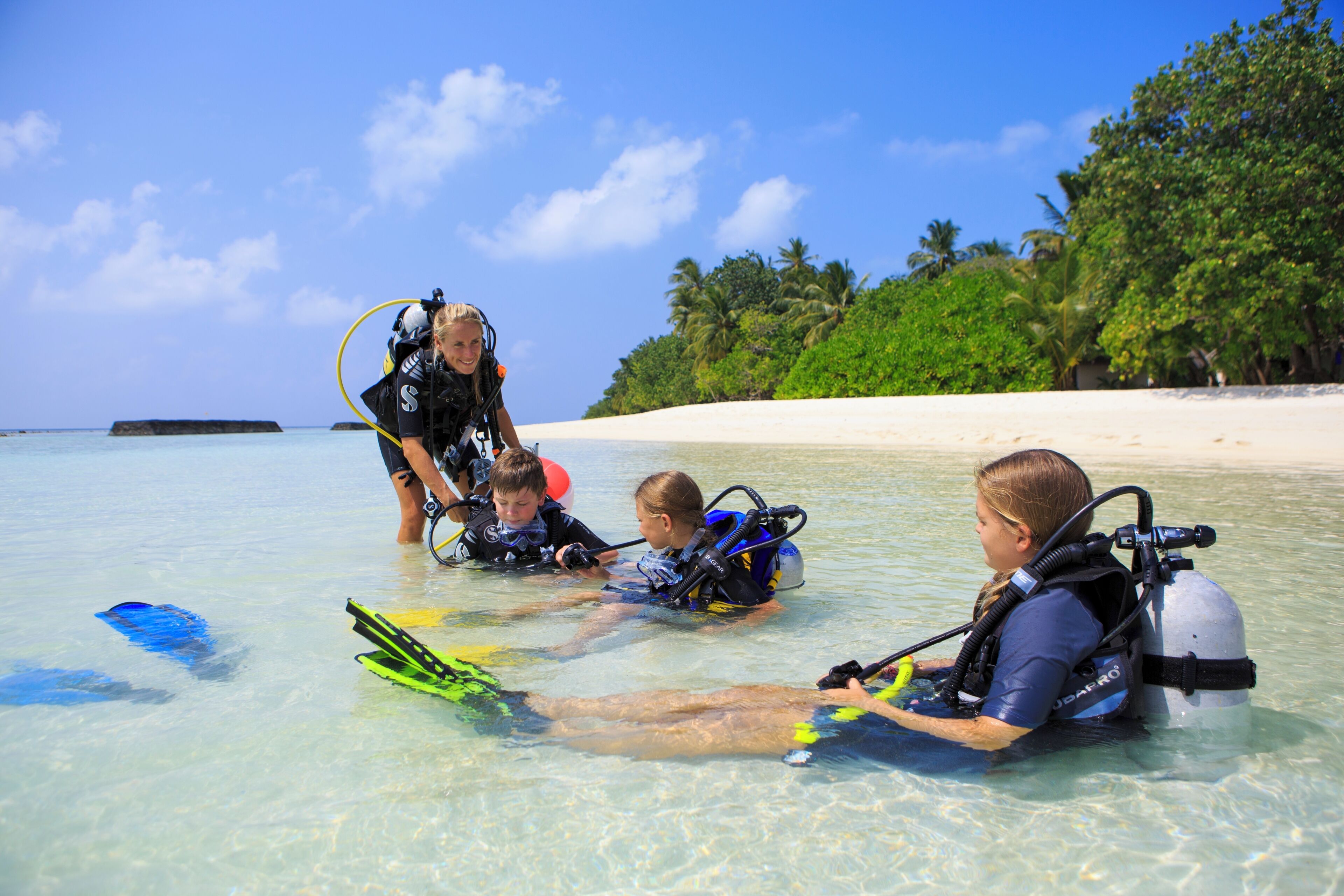 Um grupo de pessoas em um mar tropical transparente, usando equipamento de mergulho e nadadeiras, com uma praia de areia e palmeiras ao fundo.