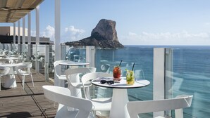 Um terraço com cadeiras e mesas brancas, uma bebida sobre uma mesa e vista para o oceano e uma ilha rochosa.