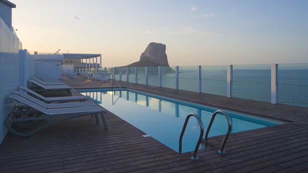 Área à beira da piscina com espreguiçadeiras, deck de madeira e vista para o mar com um conjunto de rochas.