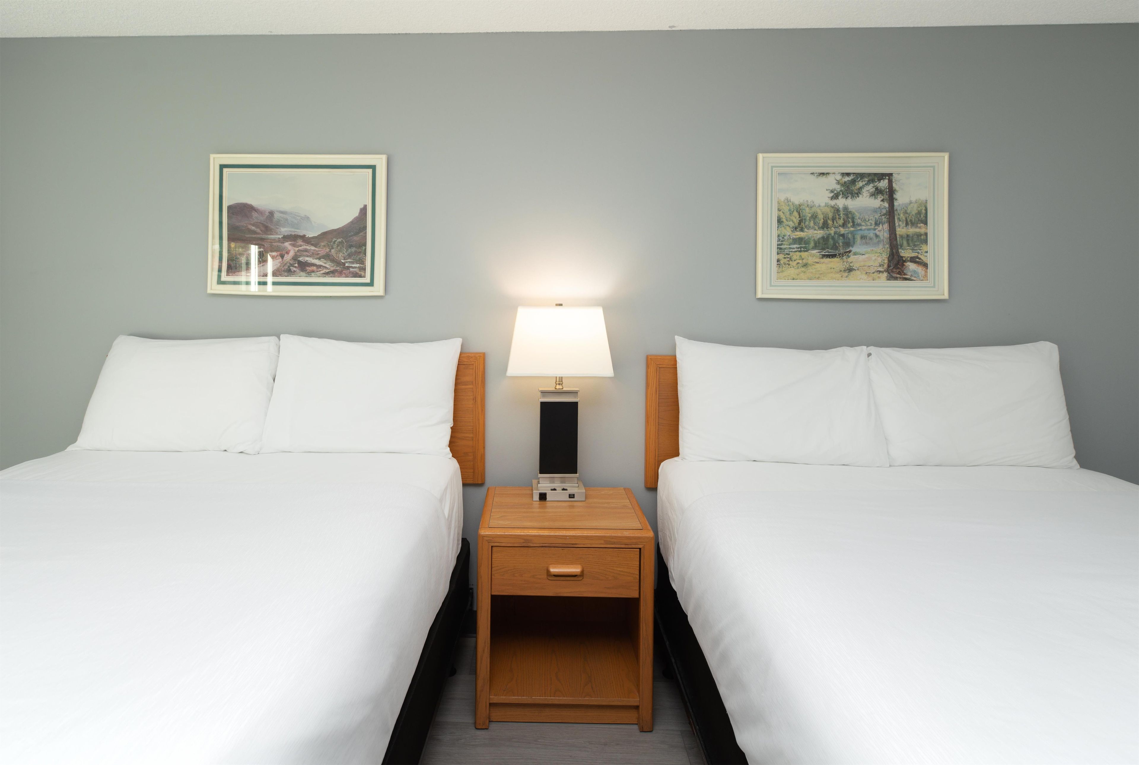 Quarto de hotel com duas camas, uma mesa de cabeceira com abajur e duas fotos emolduradas na parede.