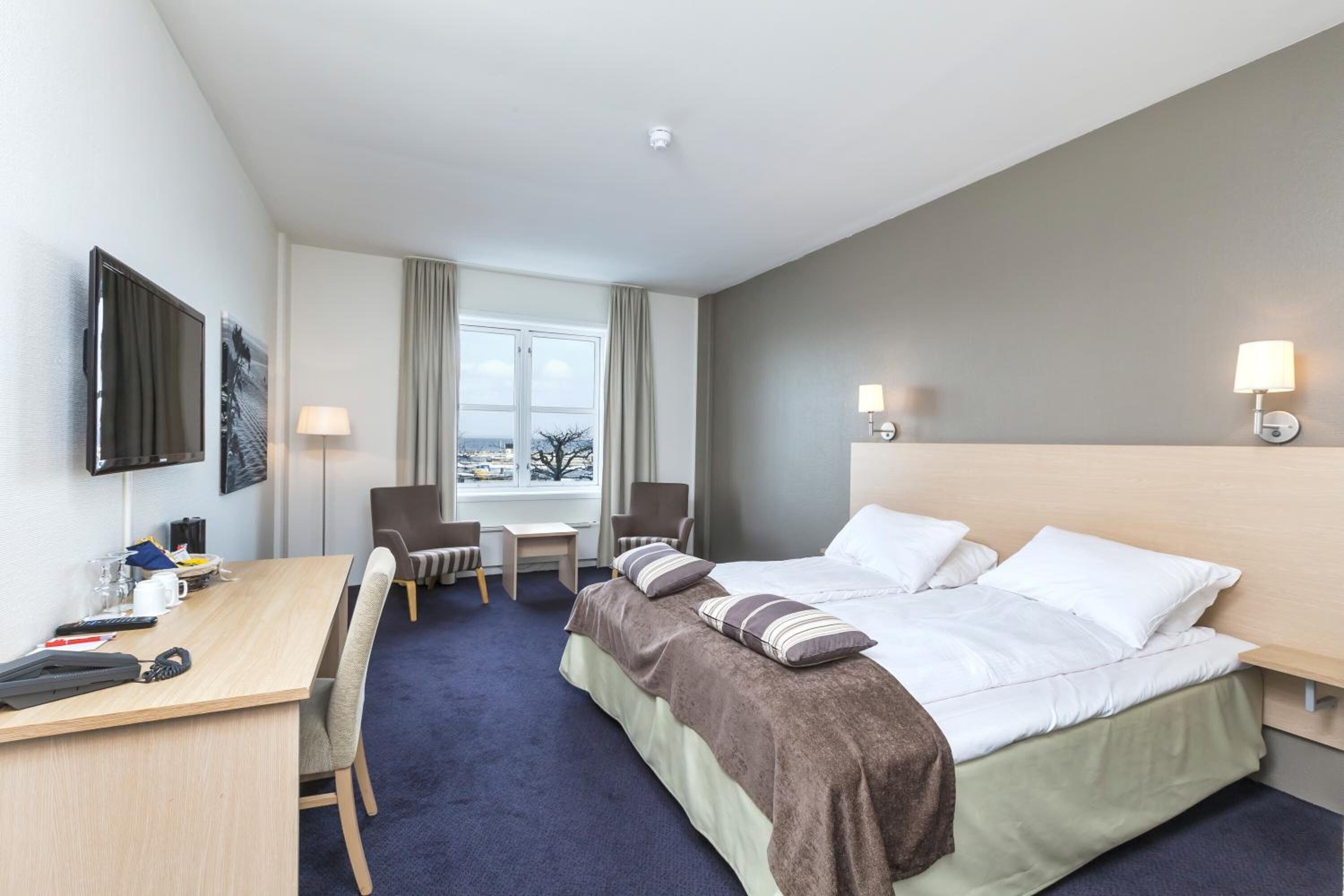 Quarto de hotel com cama, escrivaninha, cadeiras e televisão na parede.