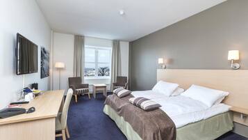 Quarto de hotel com cama, escrivaninha, cadeiras e televisão na parede.