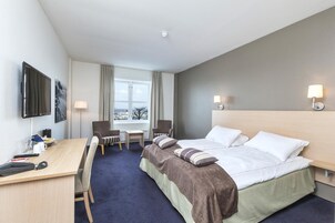 Quarto de hotel com cama, escrivaninha, cadeiras e televisão na parede.