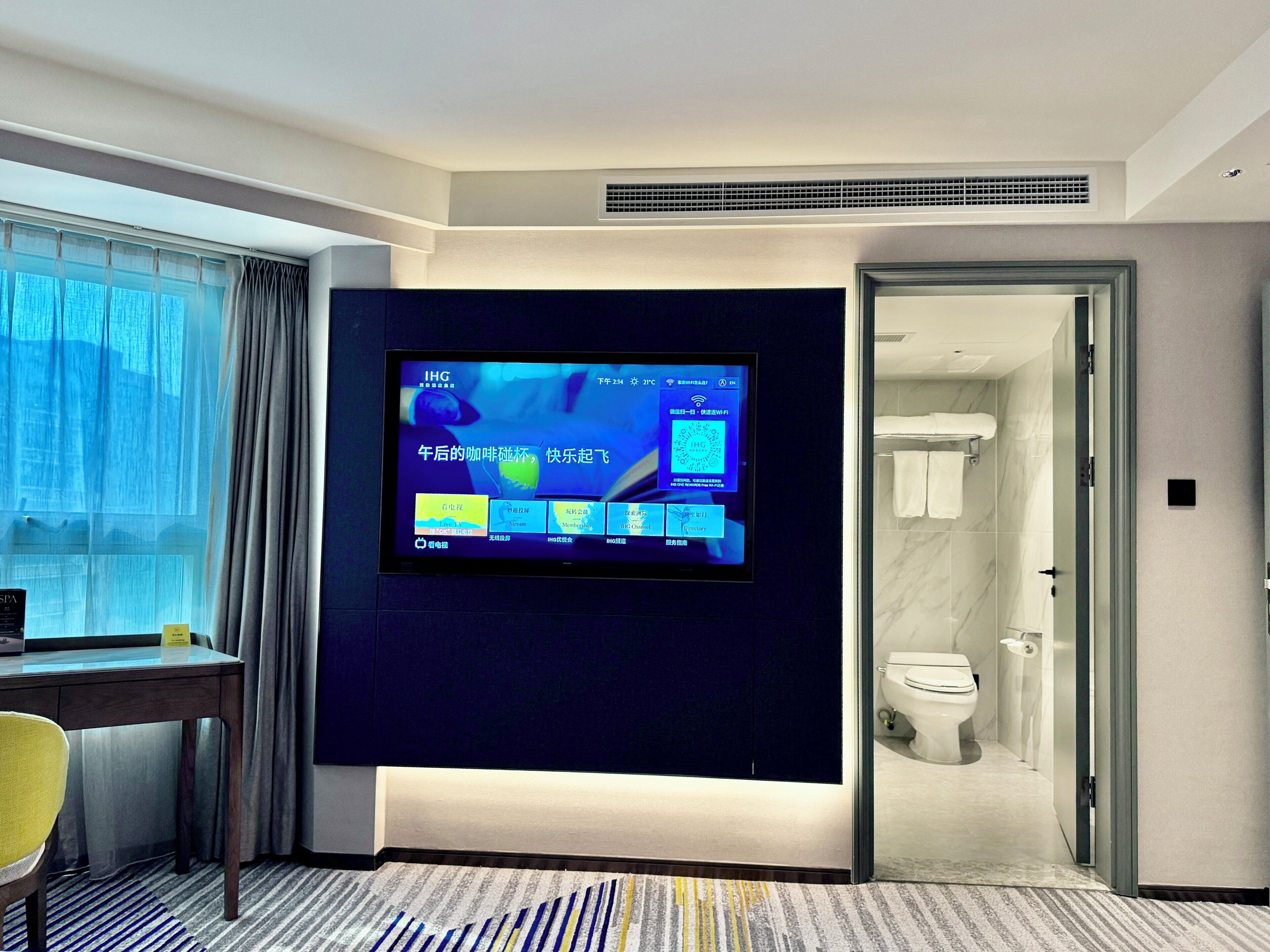 Quarto de hotel com uma televisão de tela grande fixada na parede, uma mesa com cadeira, banheiro com vaso sanitário e chuveiro, e uma janela com cortinas.