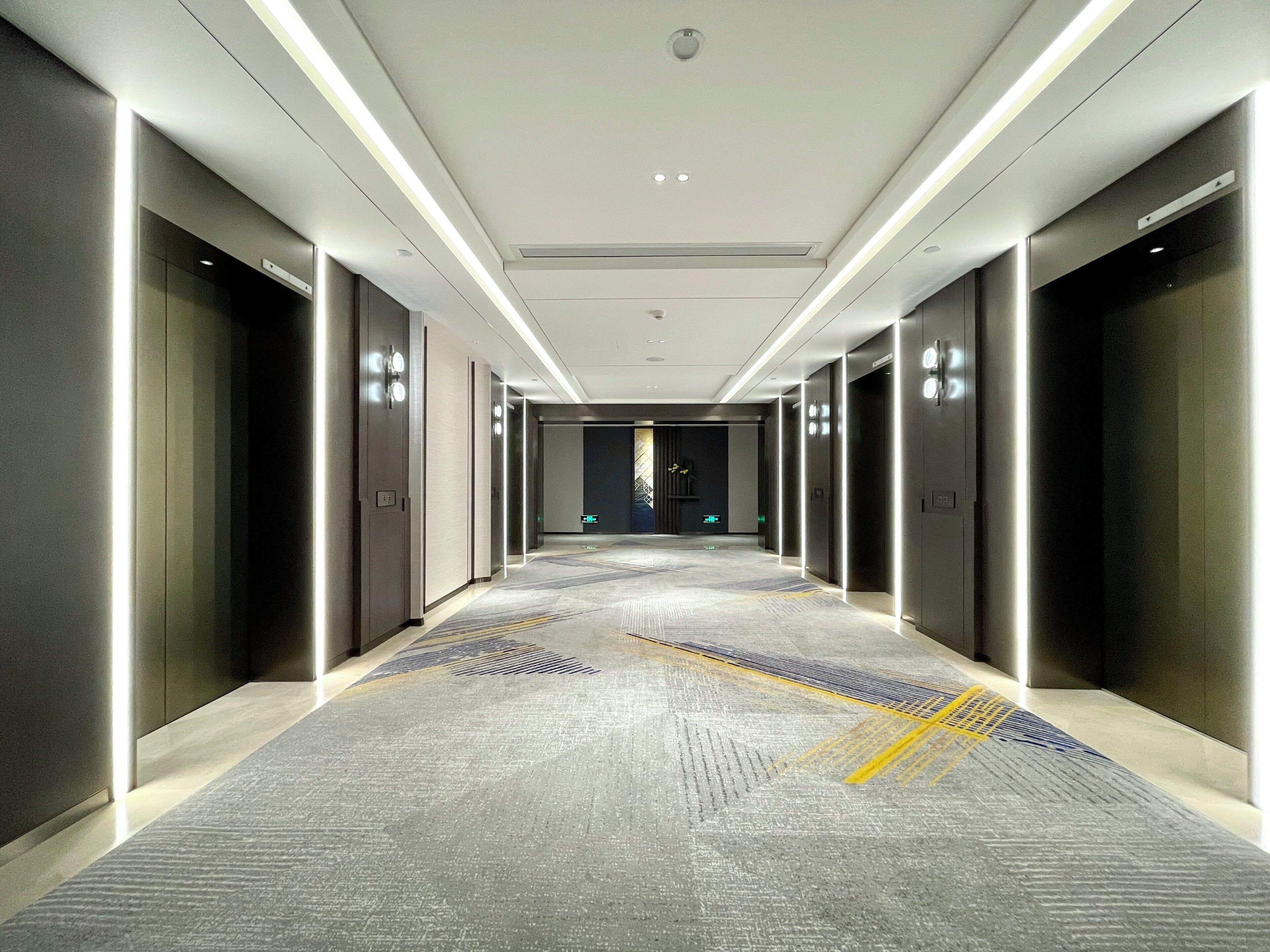 Corredor moderno de hotel com carpete estampado, iluminação embutida e uma série de portas em ambos os lados.