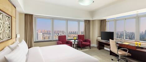 Quarto de hotel com uma cama grande, duas poltronas vermelhas, uma mesa de trabalho com computador, televisão e vista para a cidade.