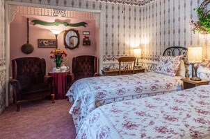 Um quarto aconchegante com duas camas, um espaço de estar com cadeiras, uma mesa pequena com um vaso de flores e um espelho na parede.
