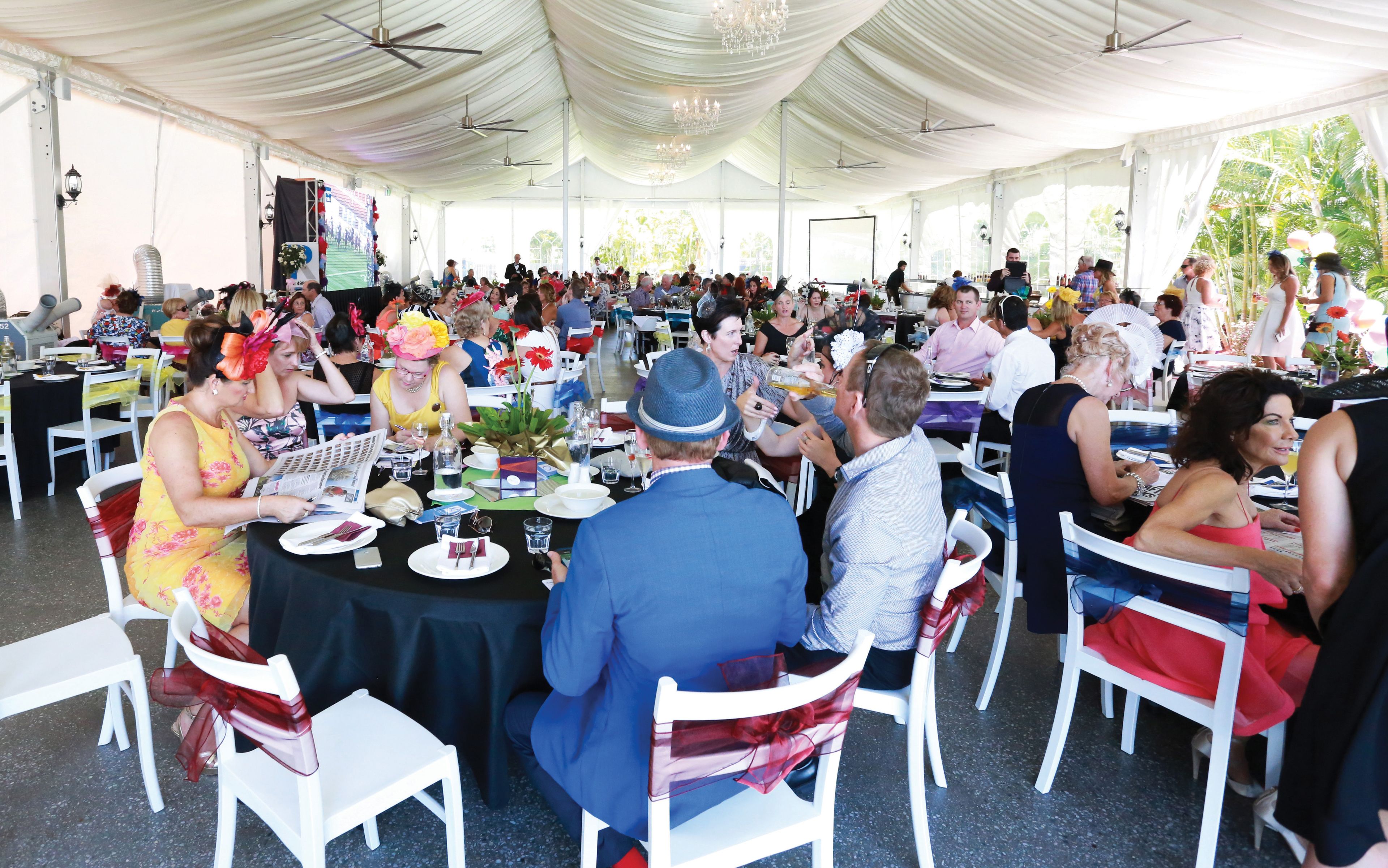 Evento ao ar livre coberto com mesas e cadeiras, participantes sentados e em pé, e um palco com uma tela ao fundo.