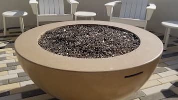 Uma área de estar circular ao ar livre com lareira, cercada por cadeiras e mesas brancas, em um pátio revestido com vista para o oceano.