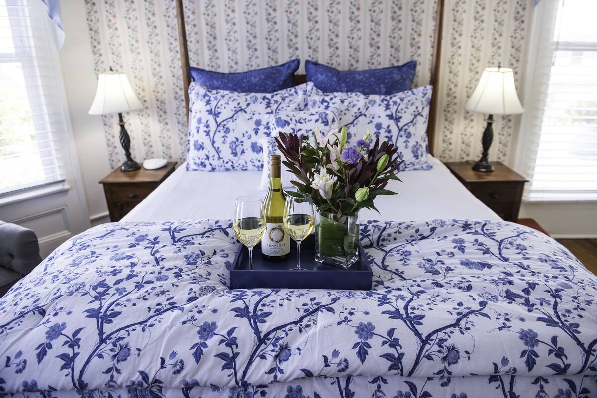 Uma cama bem arrumada com um cobreleiro floral, um tabuleiro com uma garrafa de vinho, copos e um vaso de flores.