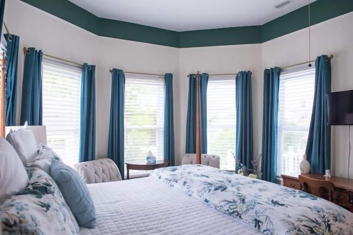 Quarto com cama grande, escrivaninha e cadeira. Possui quatro janelas com cortinas.