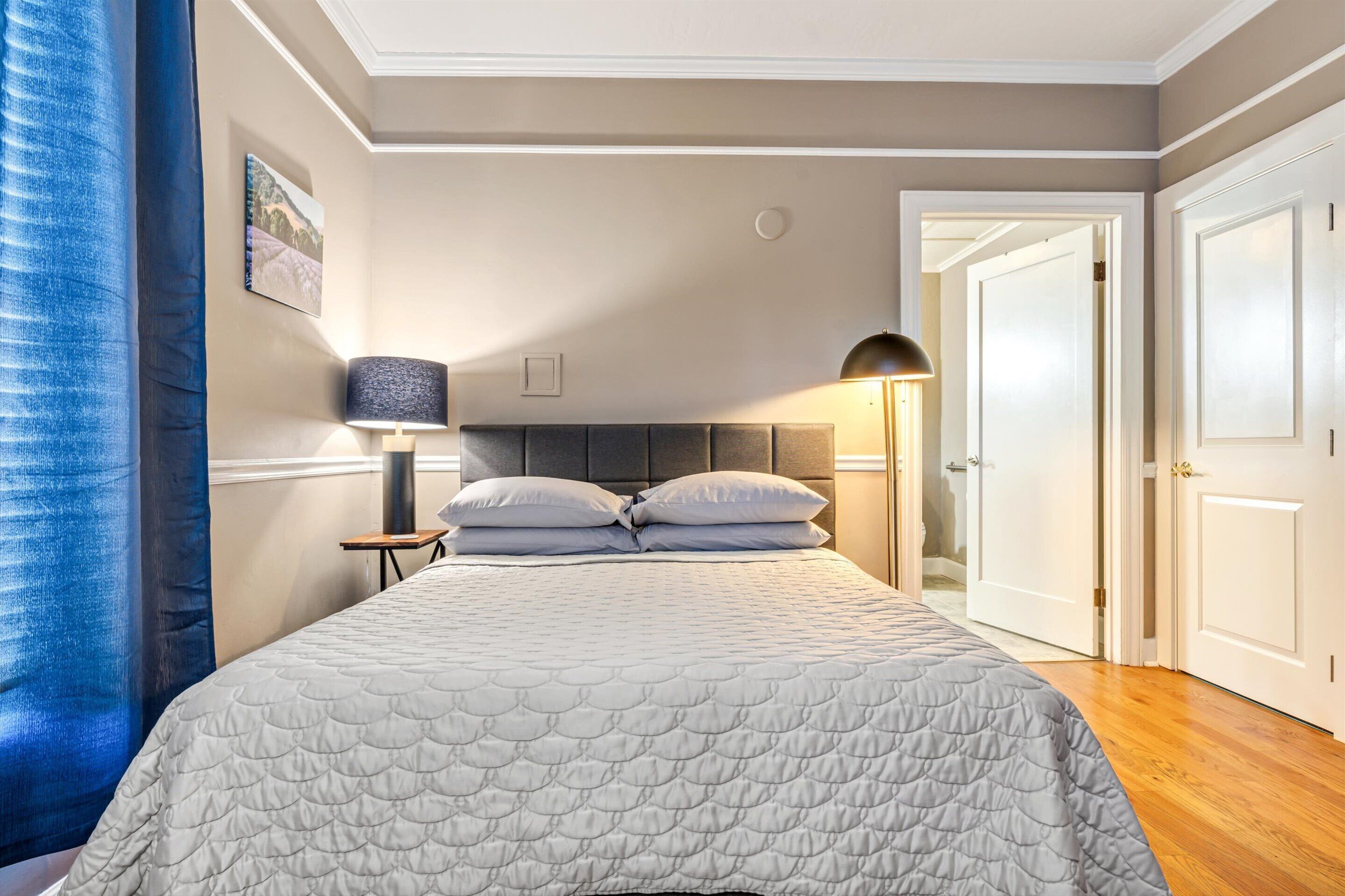 Quarto com cama, mesa de cabeceira, abajur e porta que dá para outro espaço.