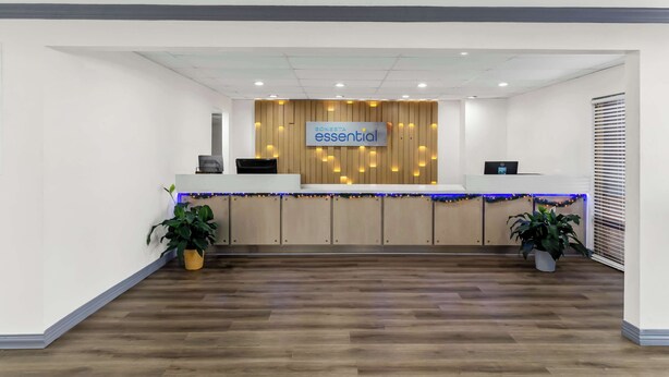 Uma área de recepção com painel de parede em madeira, uma placa com a inscrição "SONESTA essential" e duas plantas em vasos.