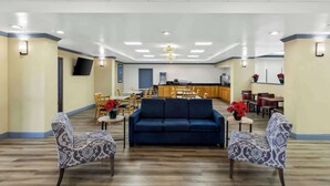 Um hall de hotel com um sofá azul, poltronas estampadas, um balcão de recepção e um espaço de refeições com mobiliário de madeira.