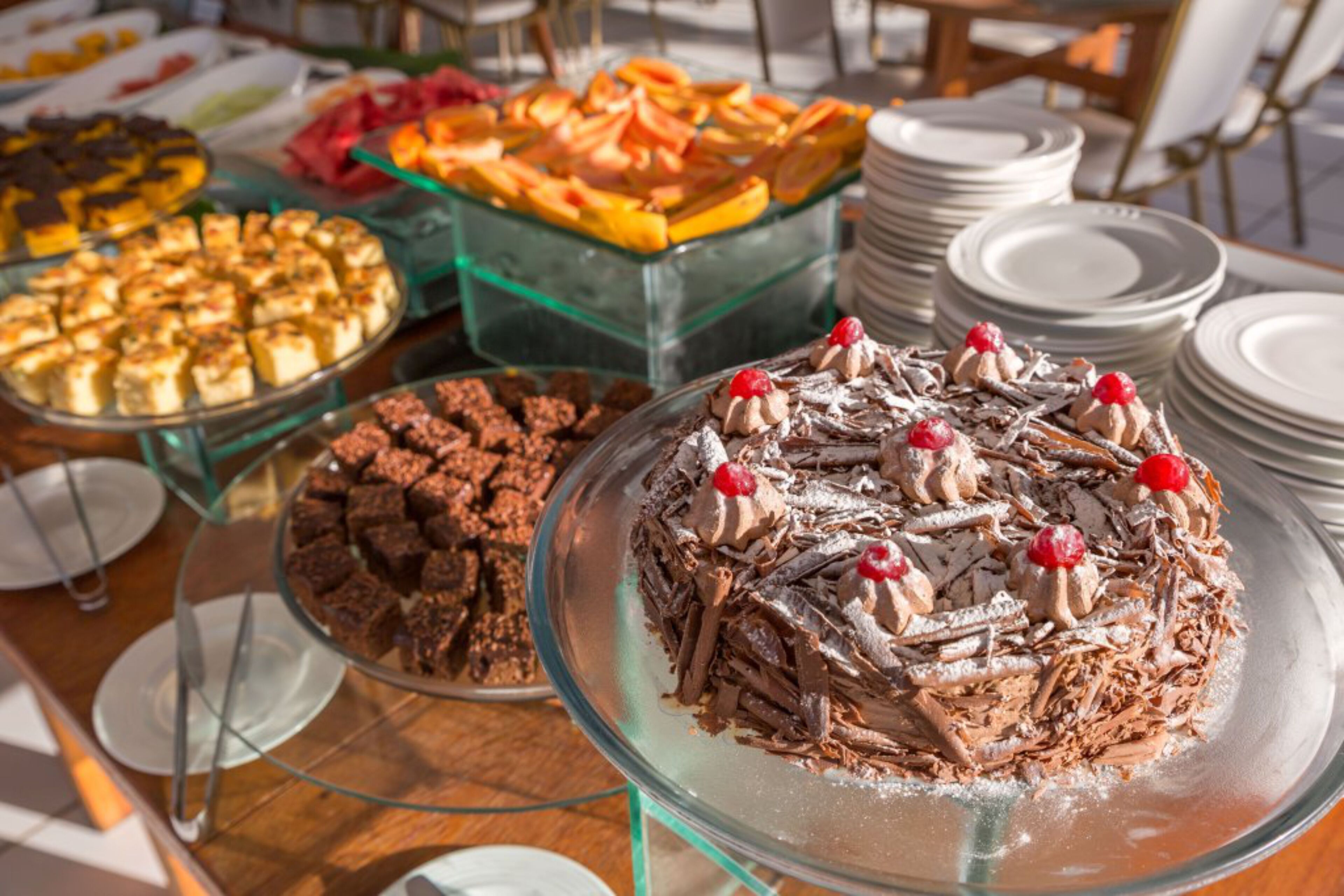 Uma mesa de buffet com diversos doces, incluindo um bolo de chocolate com chantilly e cerejas, e outras sobremesas variadas.