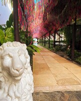 Um caminho com piso de pedra leva a um edifício, cercado por vegetação e uma cabeça de leão de pedra branca decorativa.