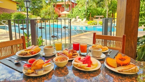 Mesa de café da manhã com diversos pratos, piscina e palmeiras ao fundo.