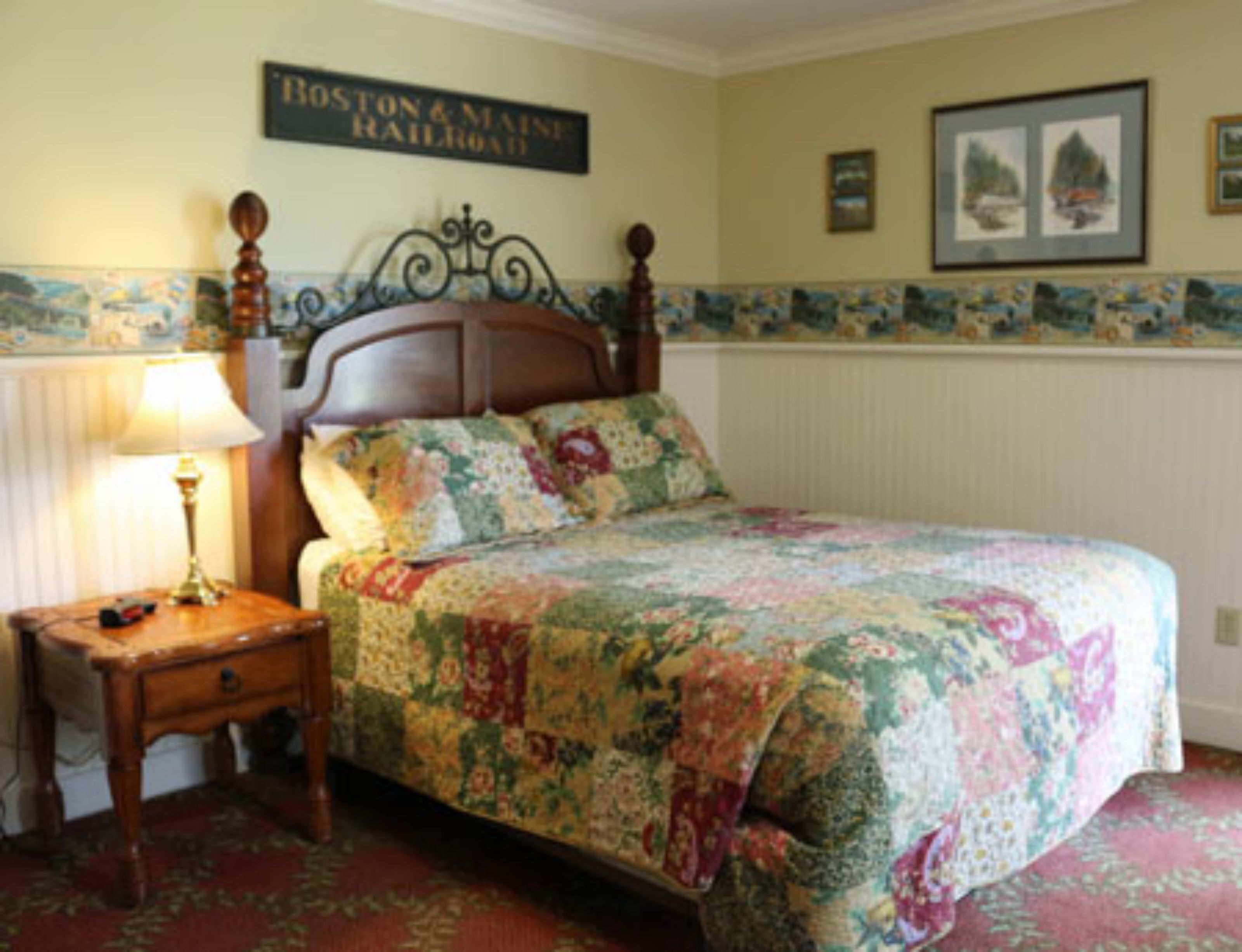 Quarto com cama de madeira, roupa de cama floral, mesa de cabeceira com abajur e um letreiro na parede com a inscrição "Boston & Maine Railroad".