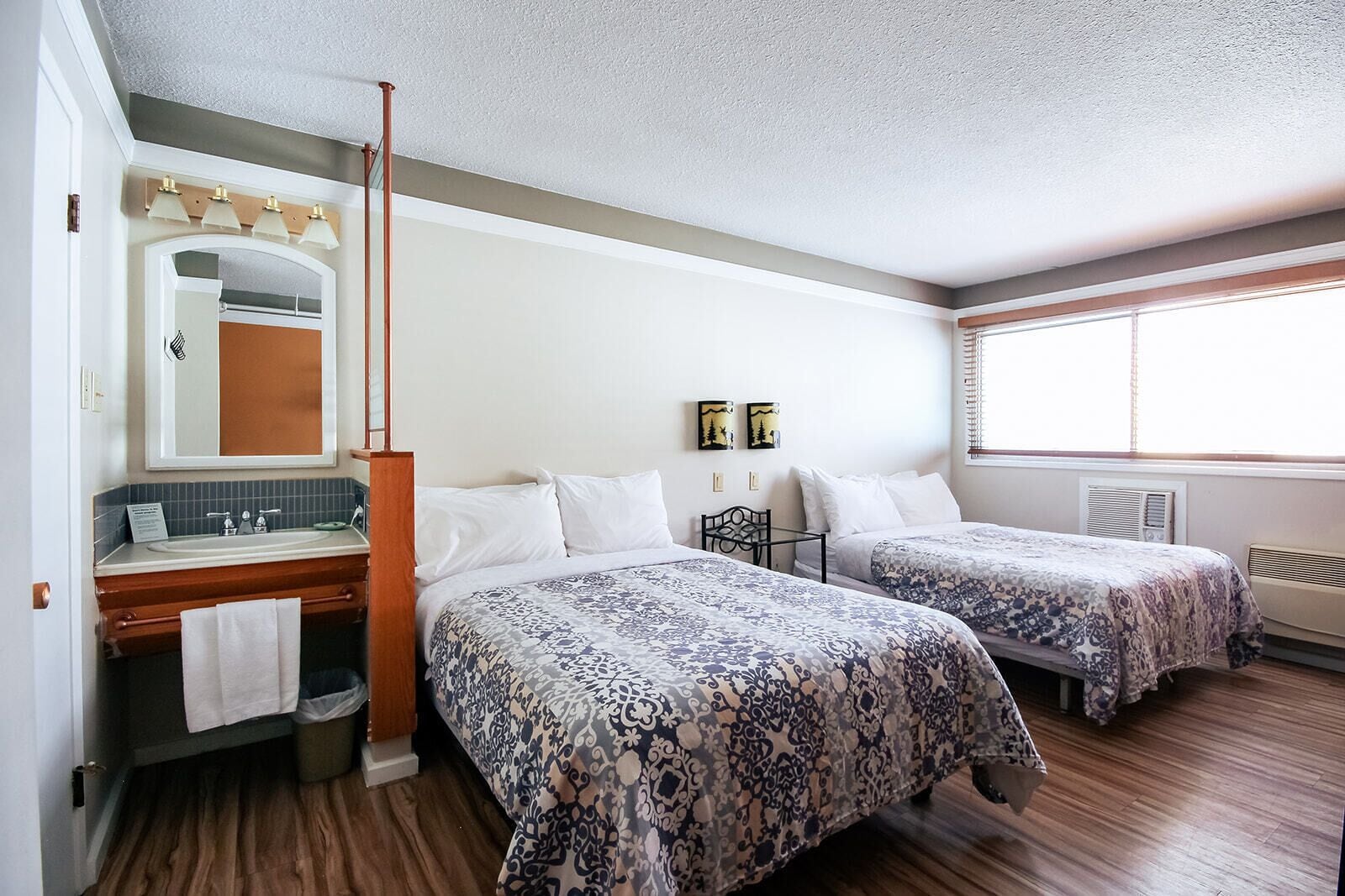 Quarto de hotel com duas camas, piso de madeira, banheiro com pia e espelho, e janela com persianas.