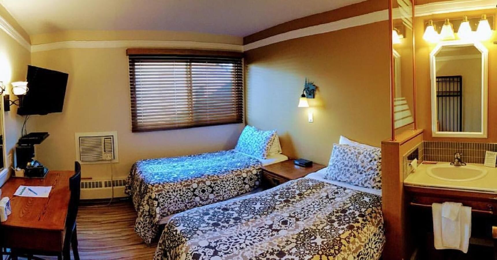 Quarto de hotel com duas camas, uma escrivaninha, uma cadeira, uma TV, uma janela com persianas, uma luminária na parede e um banheiro com pia e espelho.