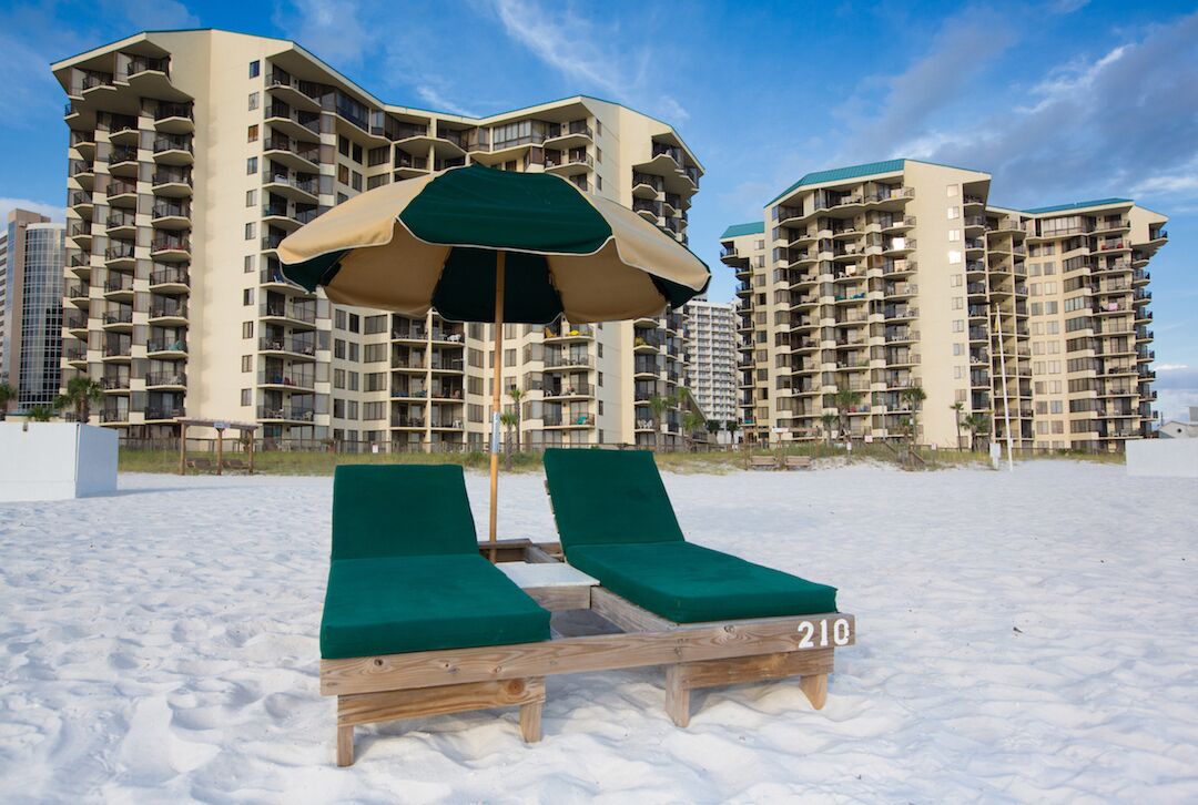 Uma praia com duas espreguiçadeiras verdes e um guarda-sol, com edifícios altos ao fundo.