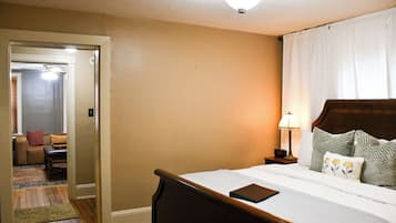 Quarto de hotel com cama, mesa de cabeceira, abajur e vista para um corredor com sofá e janela com cortinas.