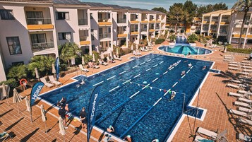 Piscina com várias pistas de natação, rodeada por espreguiçadeiras e guarda-sóis, com um complexo de edifícios ao fundo.
