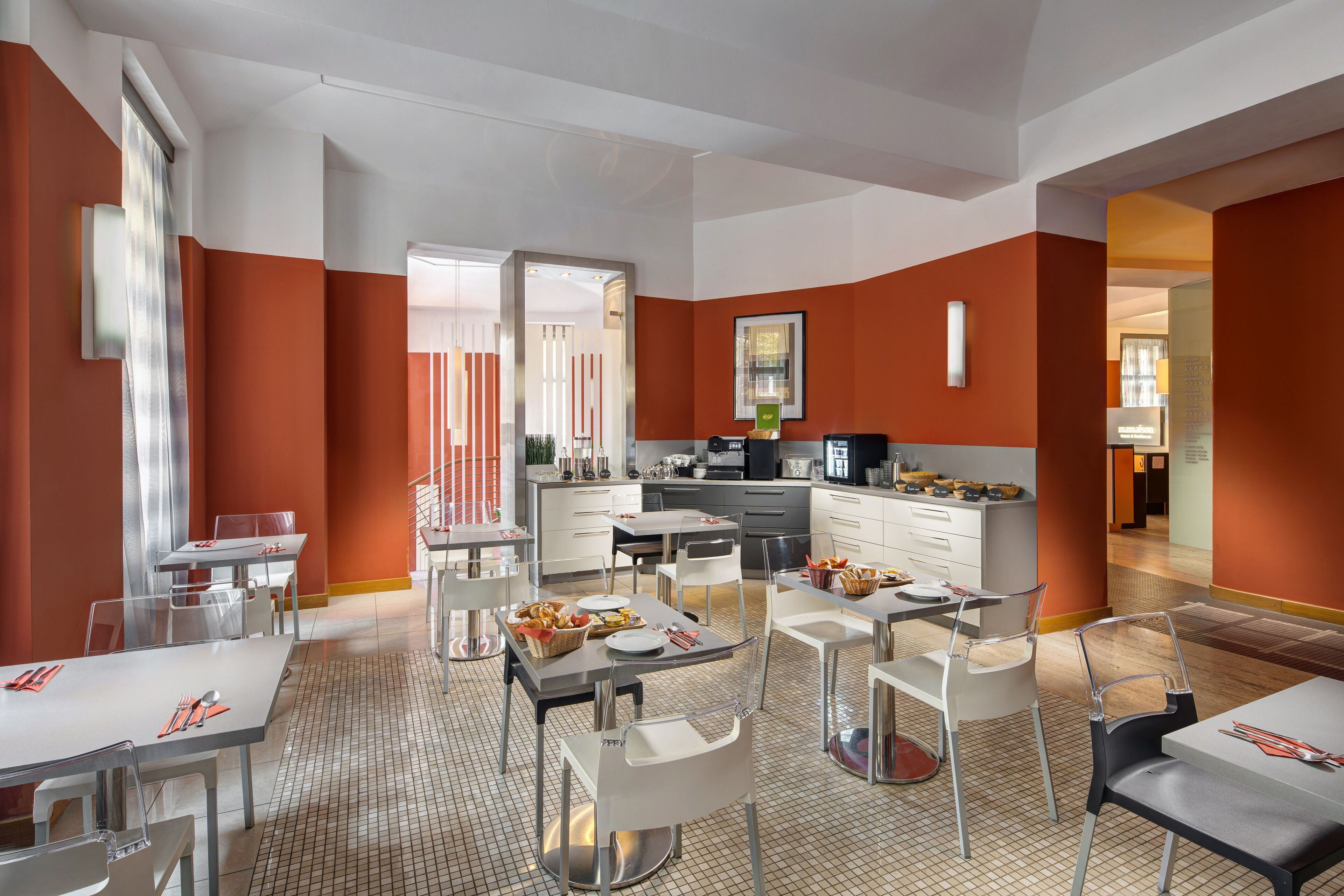 Uma sala de jantar moderna com mesas e cadeiras brancas, uma cozinha ao fundo e paredes laranja.