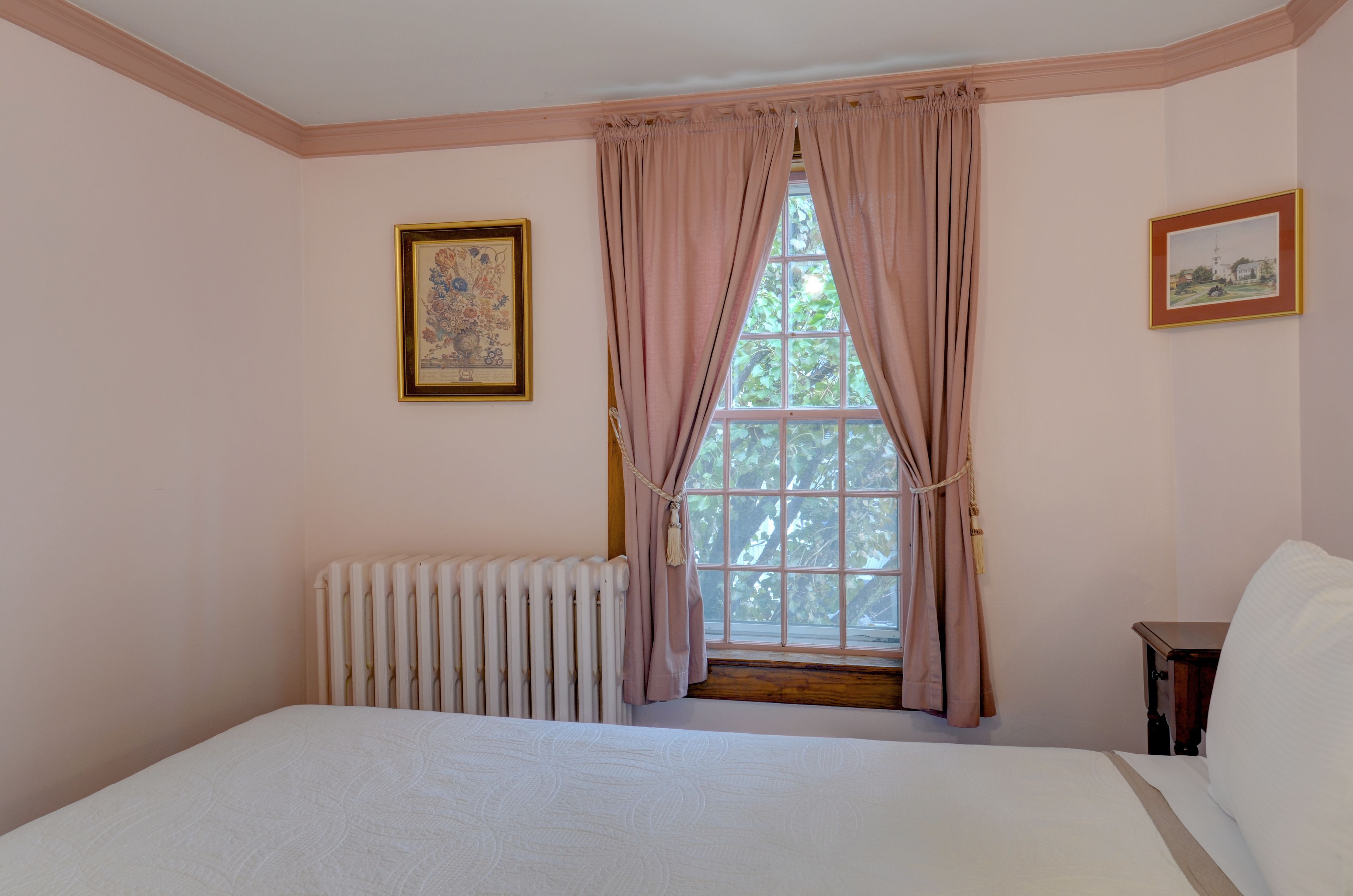 Quarto com cama grande, janela com cortinas, radiador e duas fotos emolduradas nas paredes.