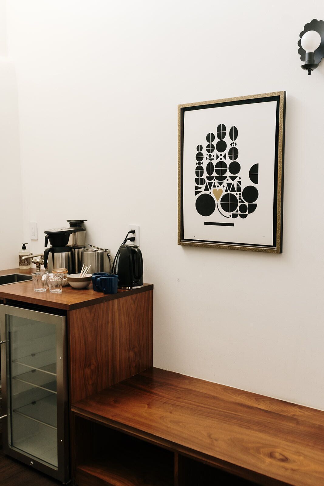 Uma cozinha compacta com cafeteira, chaleira e bule sobre a bancada, pia e um banco de madeira embaixo.