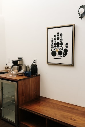 Uma cozinha compacta com cafeteira, chaleira e bule sobre a bancada, pia e um banco de madeira embaixo.
