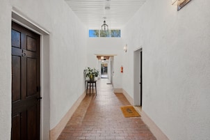 Um corredor com chão de tijolos, paredes brancas, uma porta de madeira com um número, um vaso de plantas e uma porta com extintor de incêndio.