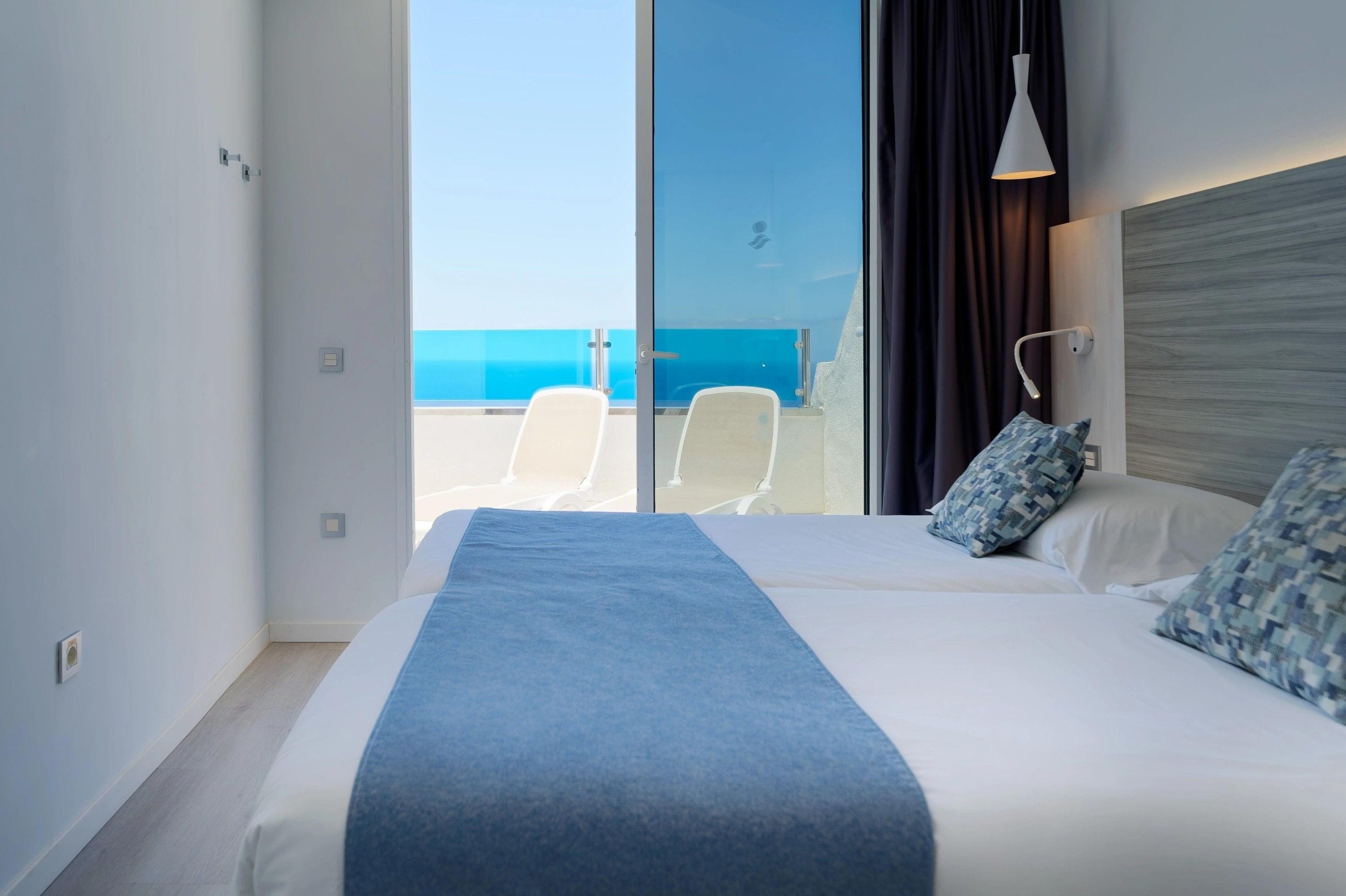 Quarto de hotel com uma cama, vista para o mar e varanda com cadeiras.