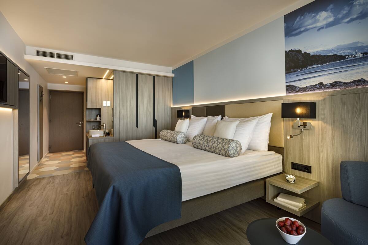 Quarto de hotel moderno com uma cama grande, mesas de cabeceira, um pequeno espaço de estar e vista para a paisagem pela janela.
