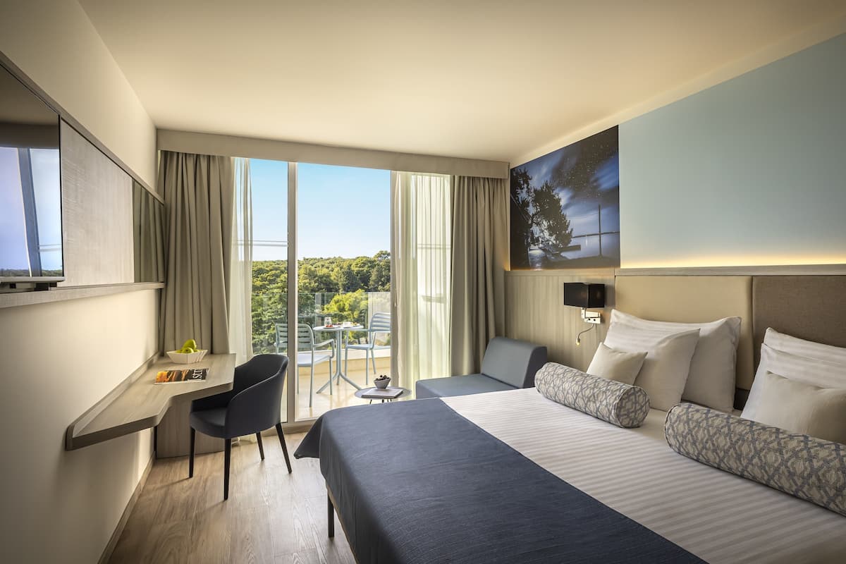 Quarto de hotel com cama, escrivaninha, cadeira e varanda com vista para a paisagem.