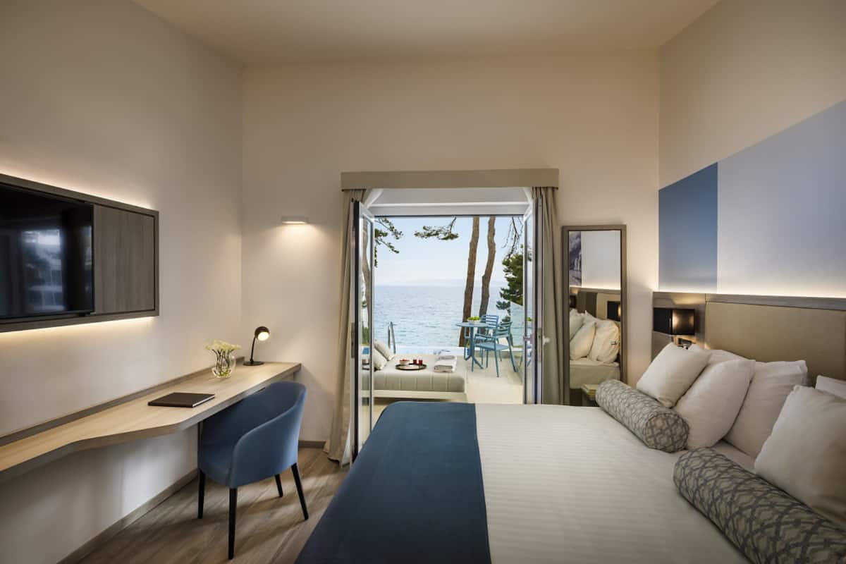 Quarto de hotel moderno com cama, escrivaninha e vista para o mar por uma porta aberta.